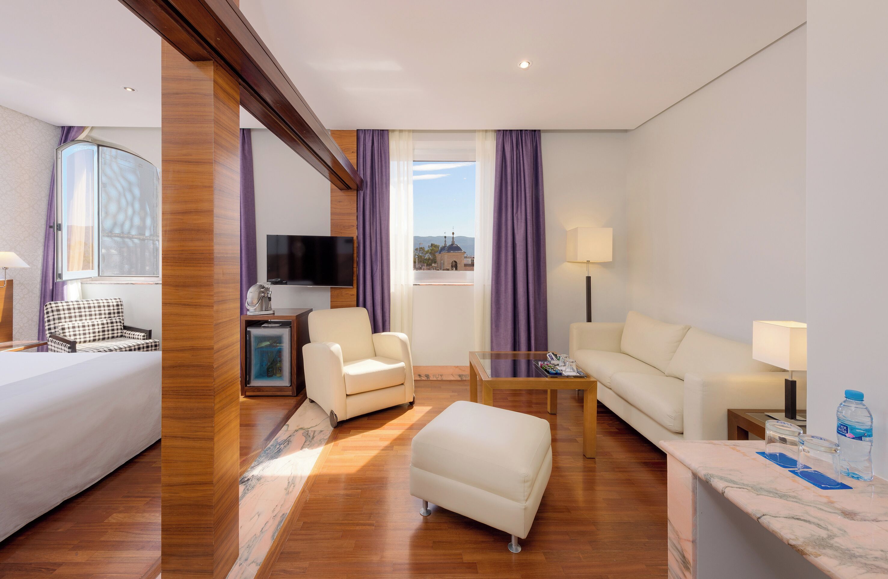 Quarto de hotel com cama, sofá, poltrona, mesa pequena, televisão e janela com cortinas.