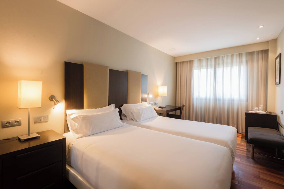 Quarto de hotel com duas camas, uma escrivaninha, uma cadeira e janela com cortinas.