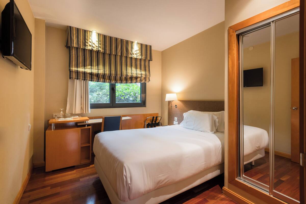 Quarto de hotel com uma cama grande, uma escrivaninha com cadeira, televisão fixada na parede e janela com cortinas.