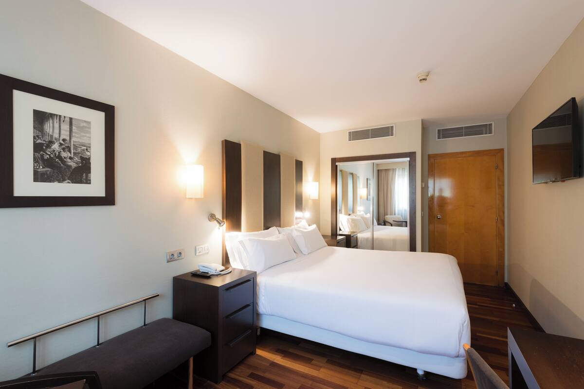 Quarto de hotel com uma cama grande, mesas de cabeceira, um banco, um quadro emoldurado, luminárias na parede, uma TV e um espelho.