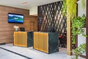 Uma área de recepção com parede de madeira, uma TV exibindo uma vista panorâmica e um balcão de recepção com tampos de mármore e uma planta à direita.