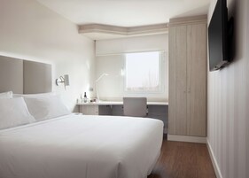 Quarto de hotel com uma cama grande, uma mesa de trabalho com cadeira, uma TV fixada na parede e uma janela com vista para o exterior.