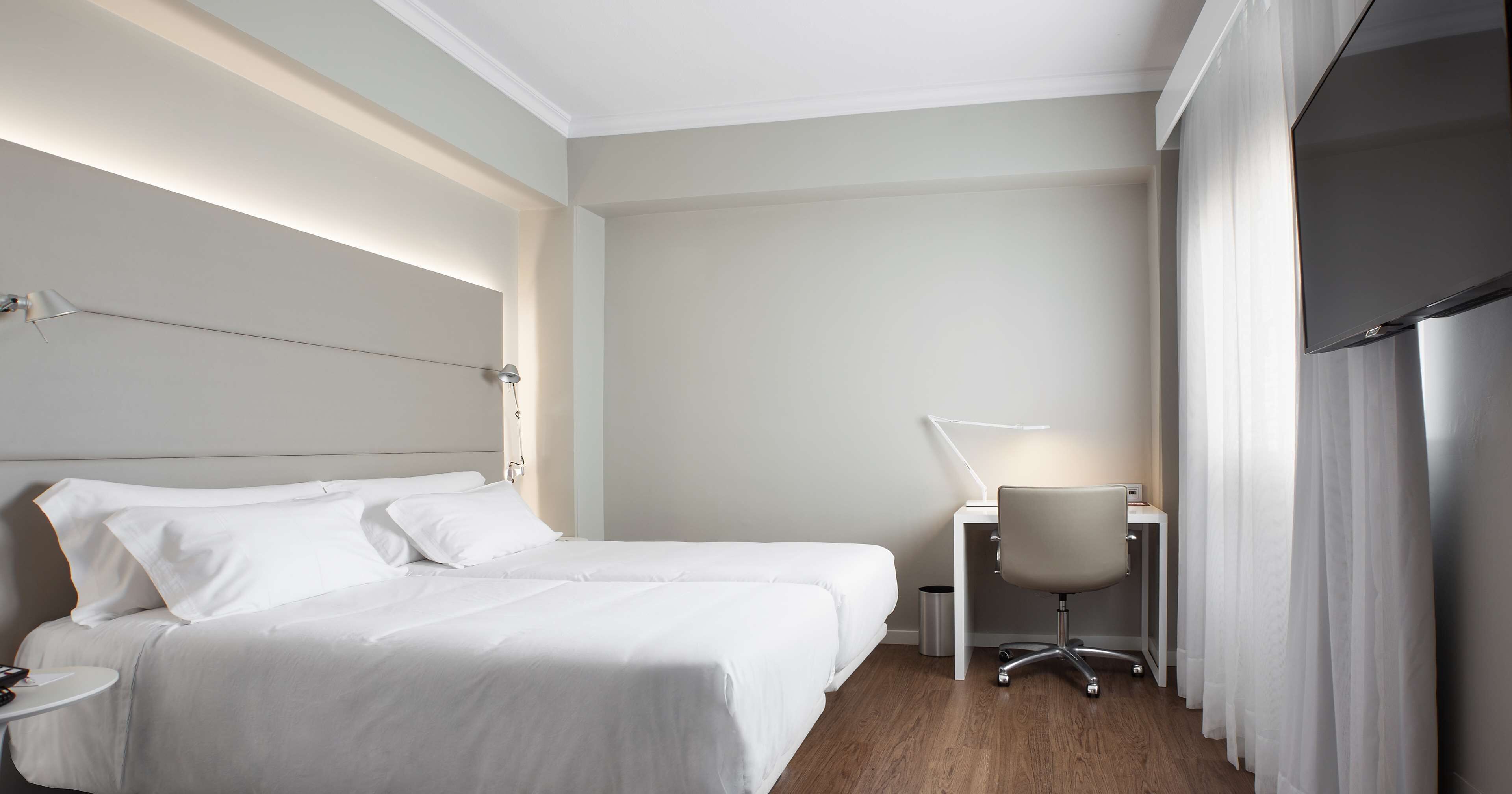 Quarto de hotel com duas camas, uma escrivaninha com cadeira, uma luminária e uma janela grande com cortinas.