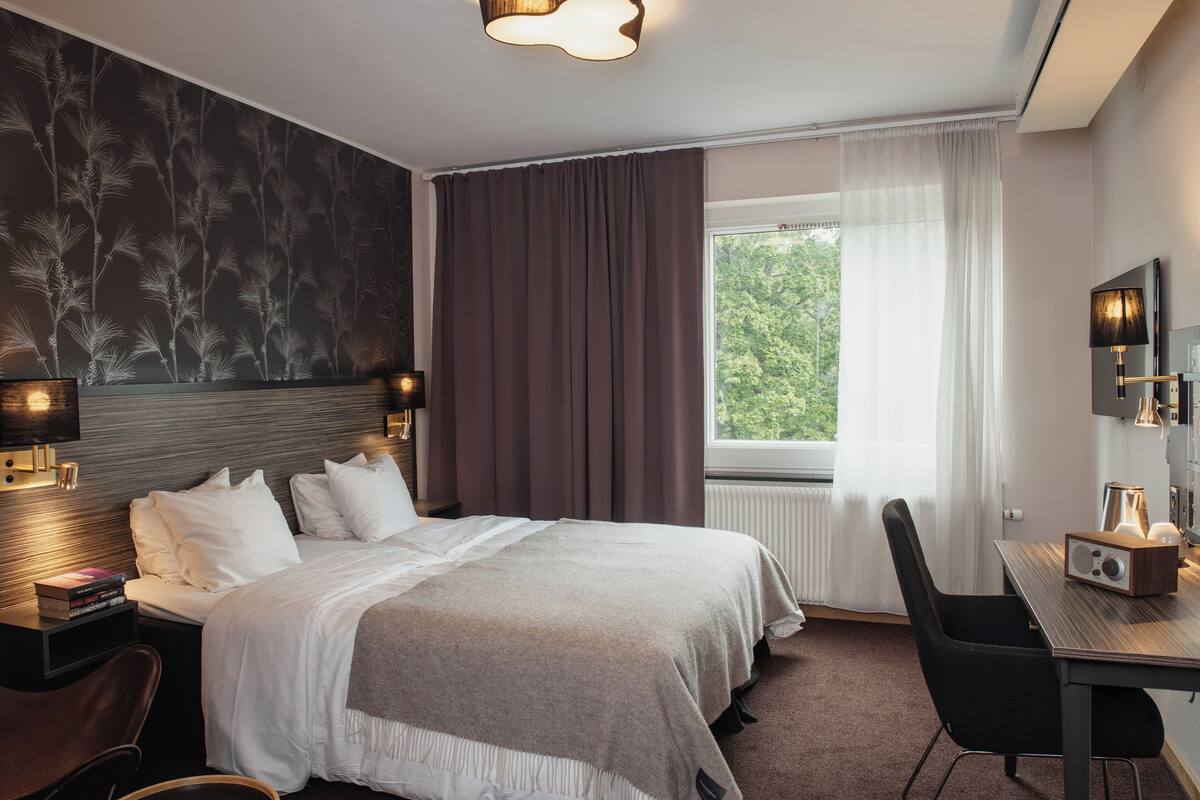Quarto de hotel com uma cama grande, uma escrivaninha com rádio, uma cadeira e uma janela com cortinas.