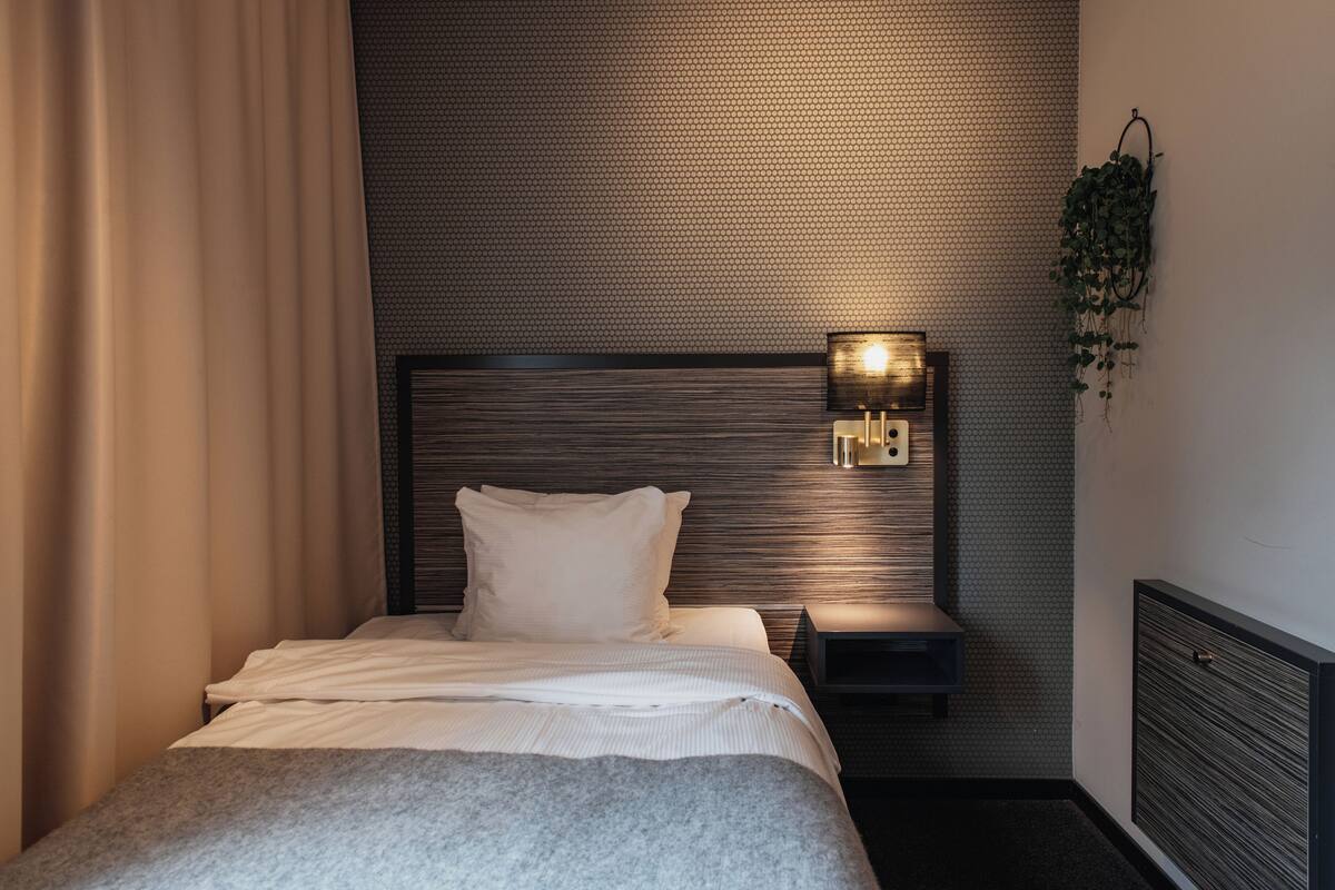 Cama bem arrumada com cabeceira, mesa de cabeceira com abajur e planta pendurada na parede.