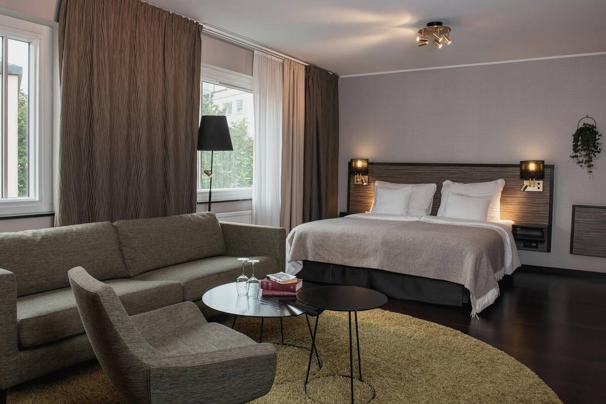 Quarto de hotel moderno com uma cama grande, um sofá, uma pequena mesa redonda e um abajur.