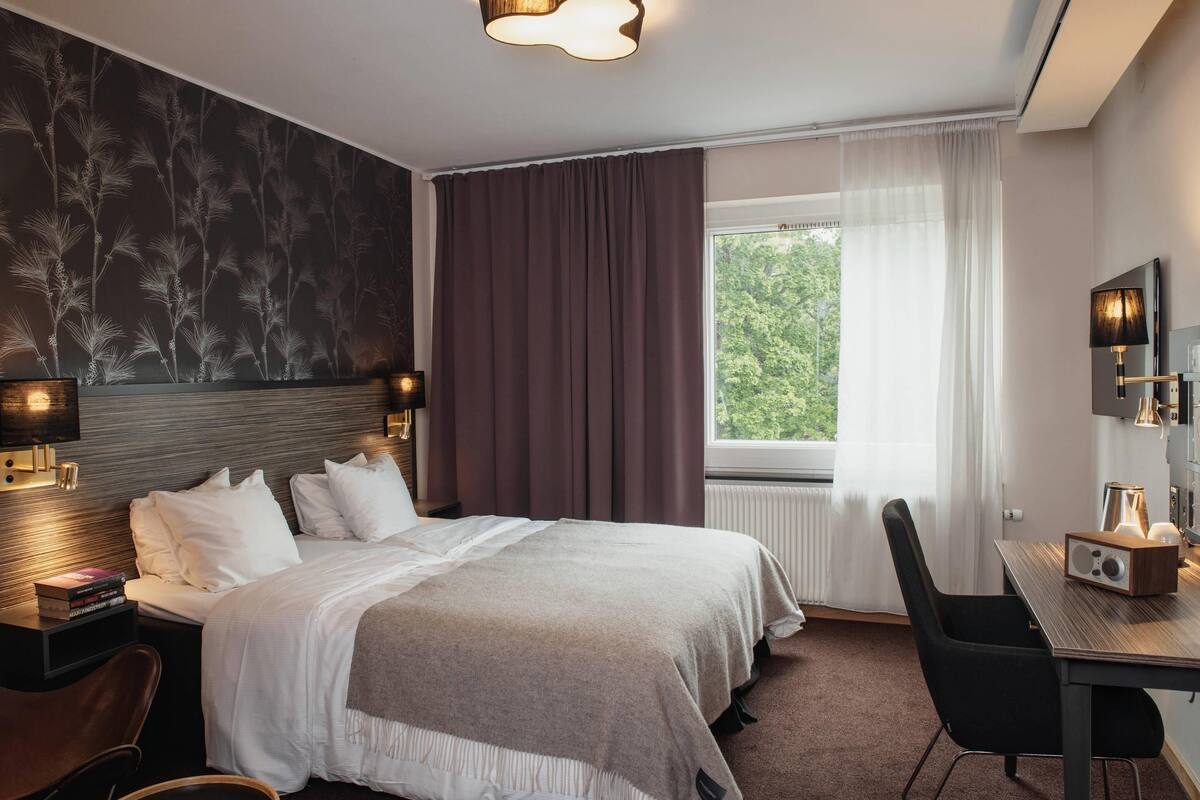Quarto de hotel com uma cama grande, uma escrivaninha com um rádio, uma cadeira e uma janela com cortinas.