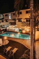 Piscina com espreguiçadeiras e guarda-sol, rodeada por um pátio de tijolos e palmeiras.