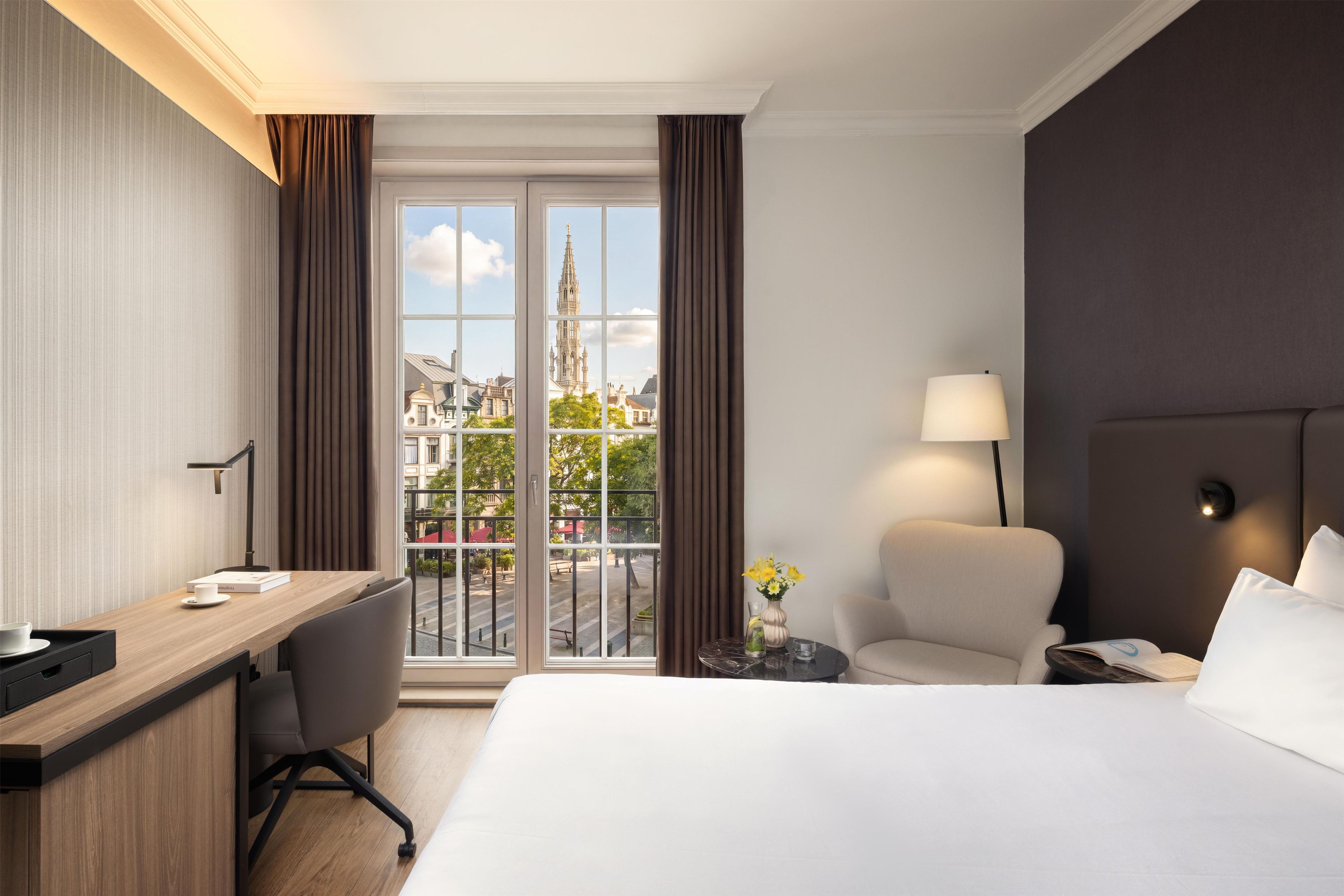 Quarto de hotel com cama, escrivaninha, cadeira, luminária e vista para o horizonte urbano através de grandes janelas.