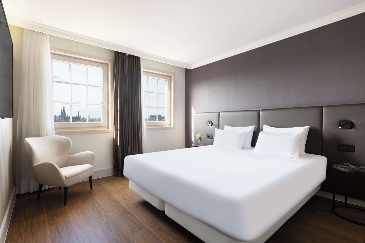 Quarto de hotel moderno com uma cama grande, uma cadeira, uma mesa de cabeceira e vista para a cidade pela janela.