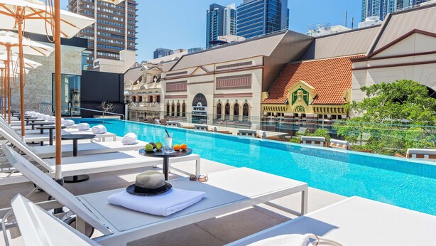 Uma área de piscina no terraço com espreguiçadeiras e guarda-sóis, uma mesa de jantar com bebida, e vista para o horizonte da cidade.