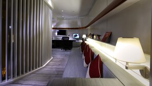 Um hall de hotel moderno com um balcão de recepção alongado, área de estar com cadeiras vermelhas, uma televisão e uma mesa com um computador.