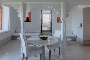 Quarto com mesa redonda, cadeiras brancas, sofá branco e vista para uma paisagem montanhosa através de uma porta aberta.
