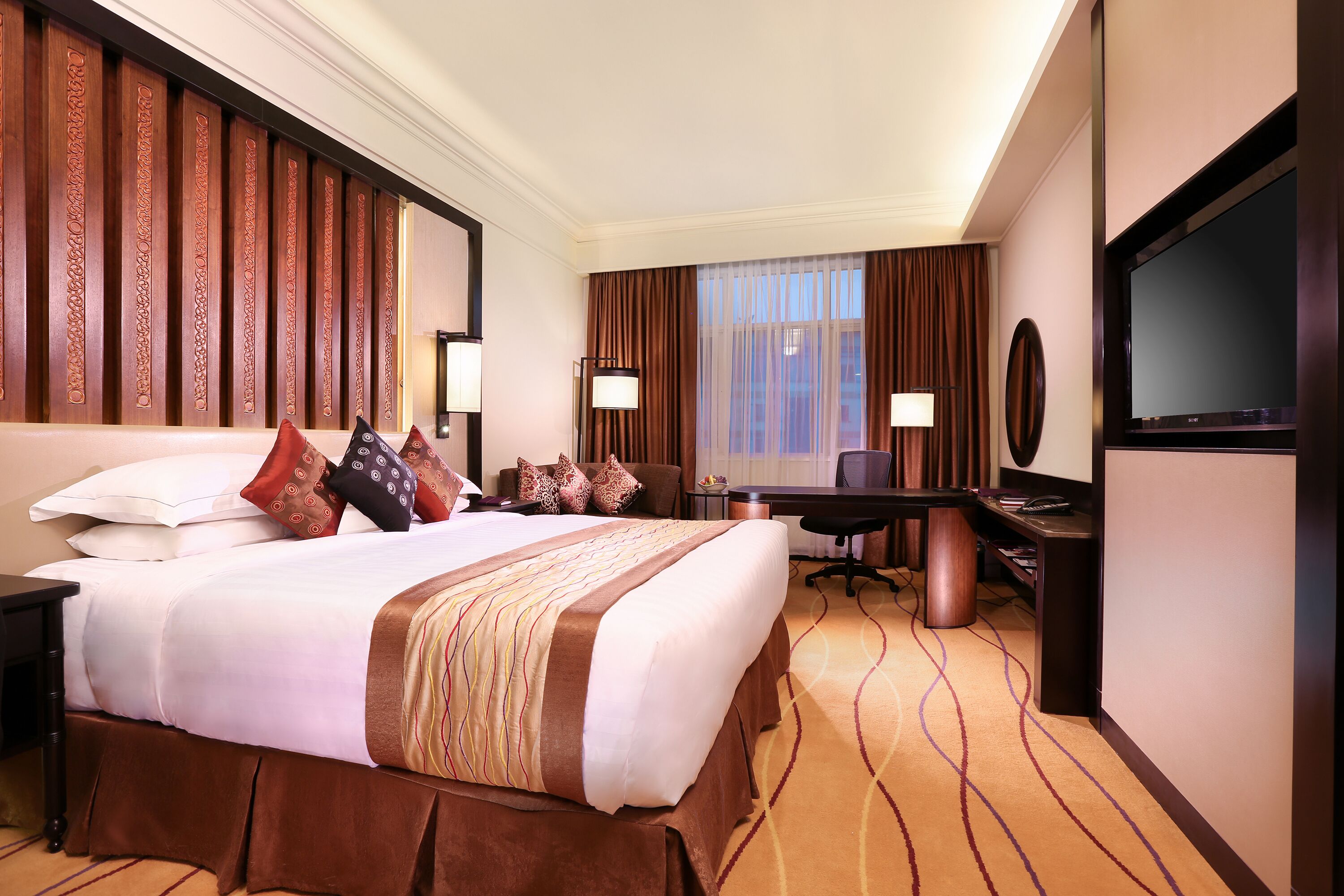 Quarto de hotel com uma cama grande, uma escrivaninha, uma televisão e uma janela com cortinas.