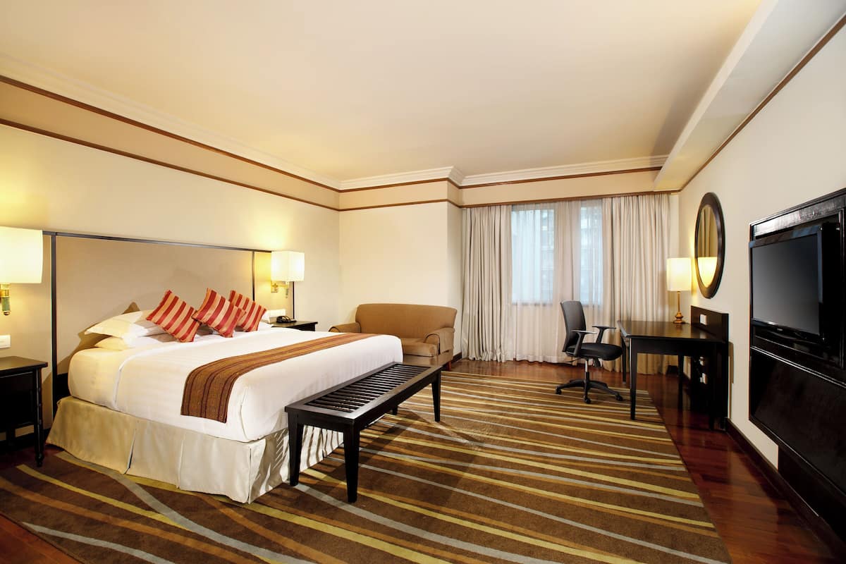 Quarto de hotel com uma cama grande, uma escrivaninha com televisão, uma cadeira e um banquinho de piano.
