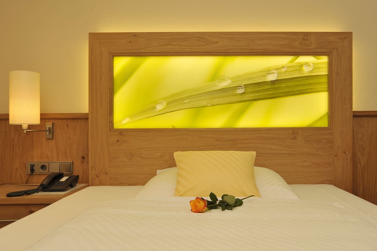Quarto de hotel com cabeceira de madeira, telefone ao lado da cama e uma cama com manta branca e um travesseiro amarelo.