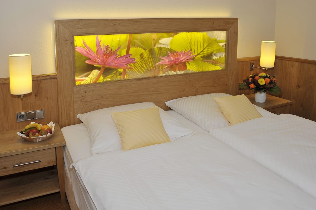 Quarto de hotel com cabeceira de madeira, duas luminárias de apoio, uma cesta de frutas e um vaso com flores.