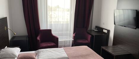 Quarto de hotel com cama, duas poltronas, escrivaninha, televisão e janela com cortinas.