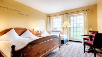 Quarto de hotel com cama de madeira, uma mesinha com cadeira, janela com cortinas e vista para o verde lá fora.