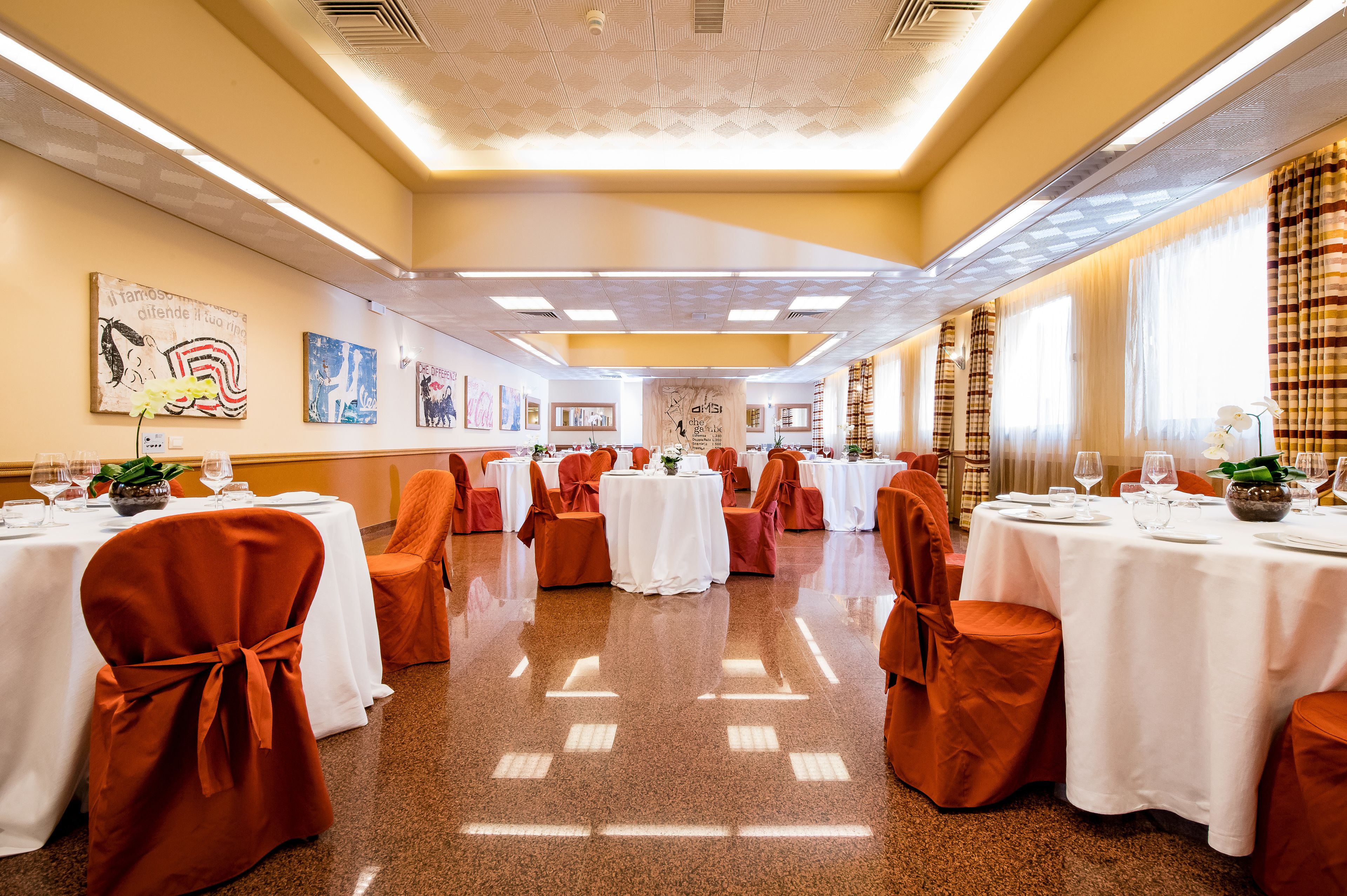 Uma sala de banquetes com várias mesas redondas, cada uma com toalhas brancas e cadeiras laranja. O espaço possui piso polido, obras de arte nas paredes e janelas amplas com cortinas.