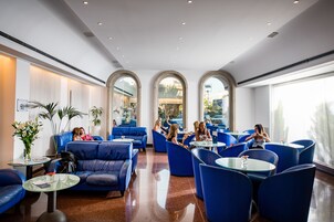 Um lounge moderno com assentos azuis e mesas de vidro, janelas amplas e plantas em ambiente interno.
