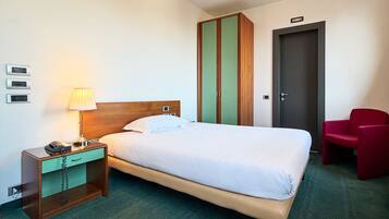 Quarto de hotel com cama, mesa de cabeceira, luminária e uma cadeira vermelha.
