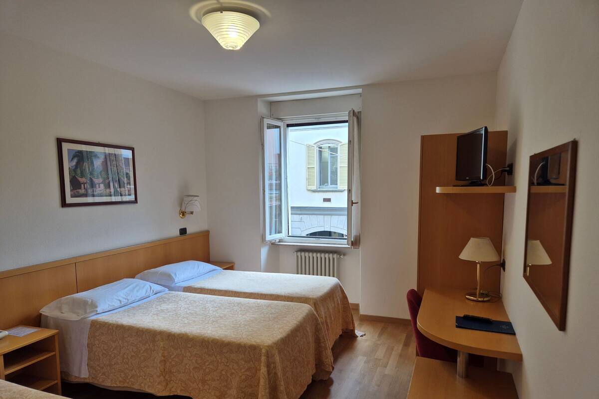 Quarto de hotel com duas camas, uma janela, uma televisão em uma prateleira e uma luminária em uma mesinha de cabeceira.