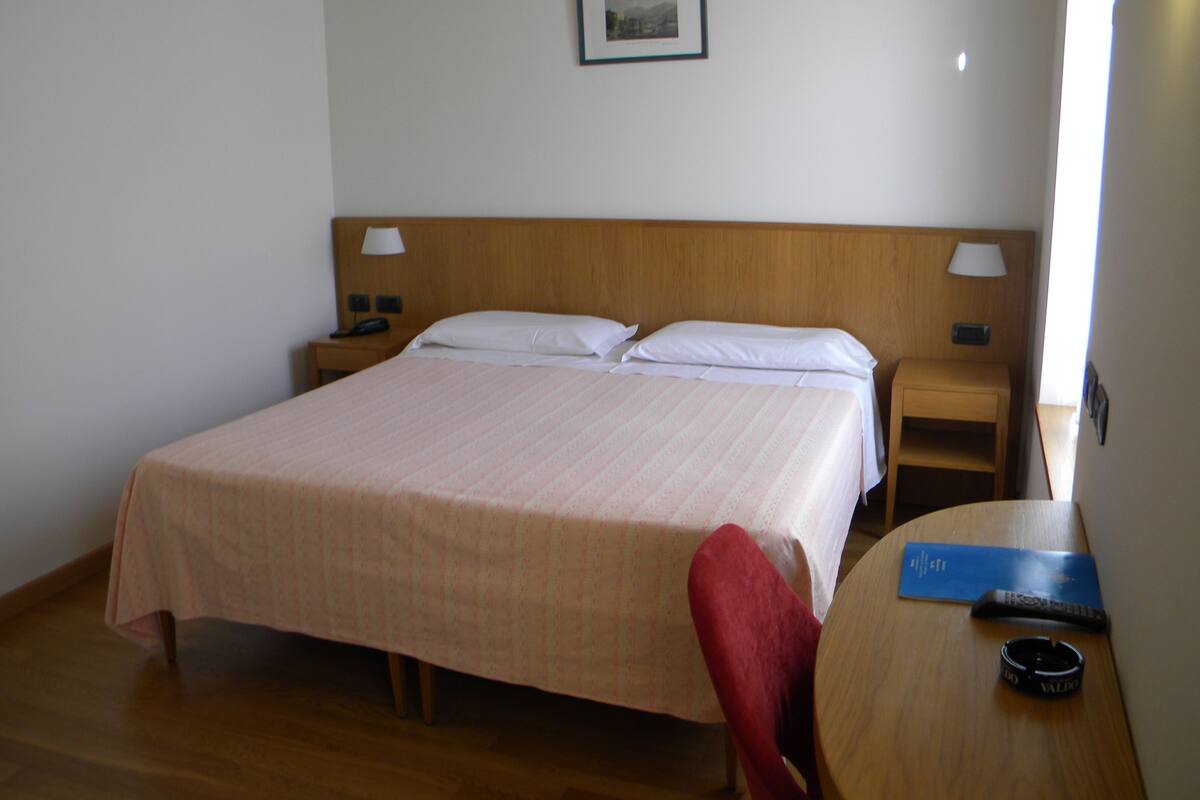 Quarto de hotel com uma cama grande, duas mesas de cabeceira, um quadro na parede e uma mesinha com um controle remoto e um livro.