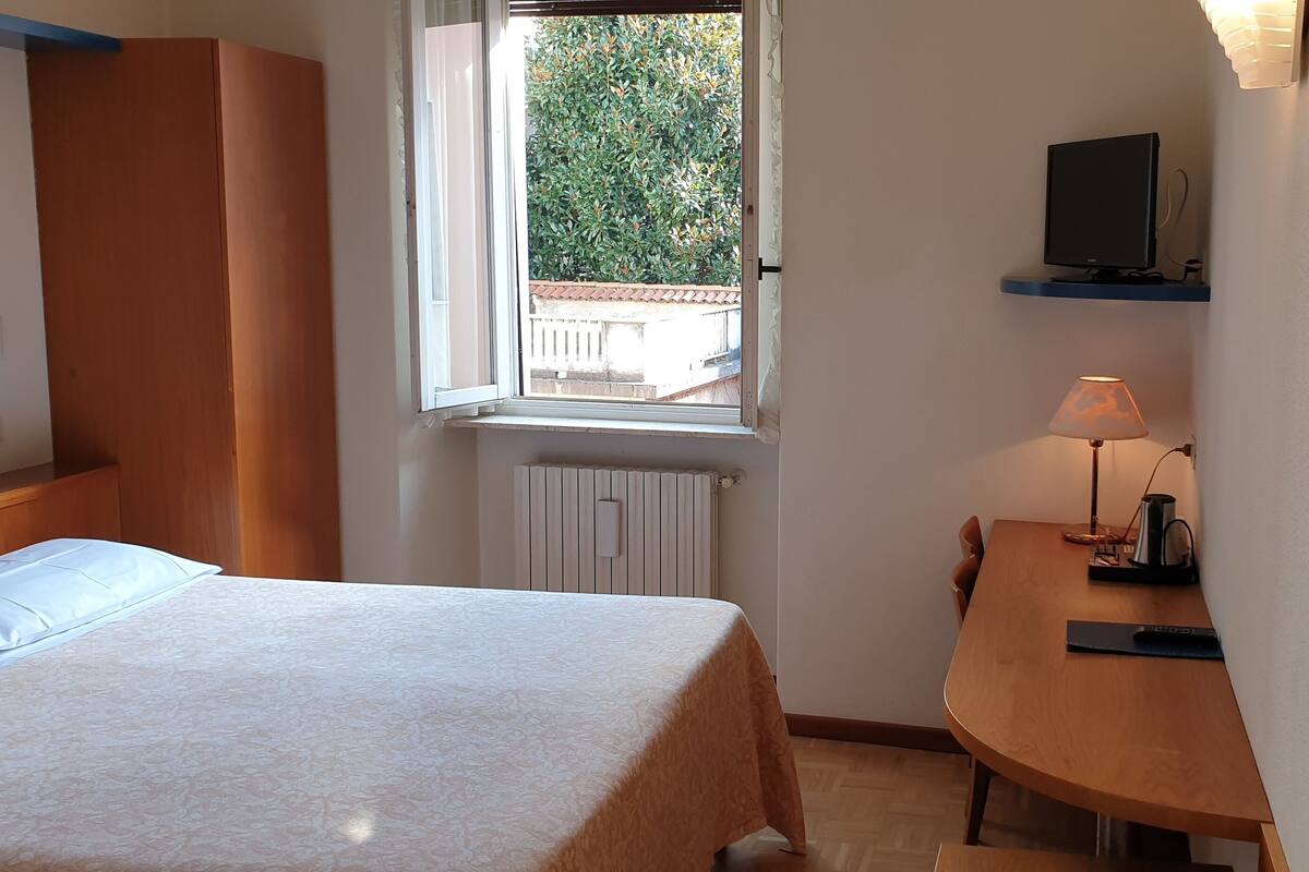 Quarto com cama, janela, escrivaninha com luminária e TV em uma prateleira.