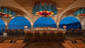 Uma área de bar com vista para o mar, apresentando teto de madeira, luminárias azuis pendentes e diversos bancos e mesas de bar.