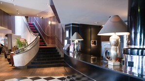 Um lobby de hotel moderno com piso xadrez, um balcão de recepção curvo, uma escadaria com degraus vermelhos e uma máquina de café.