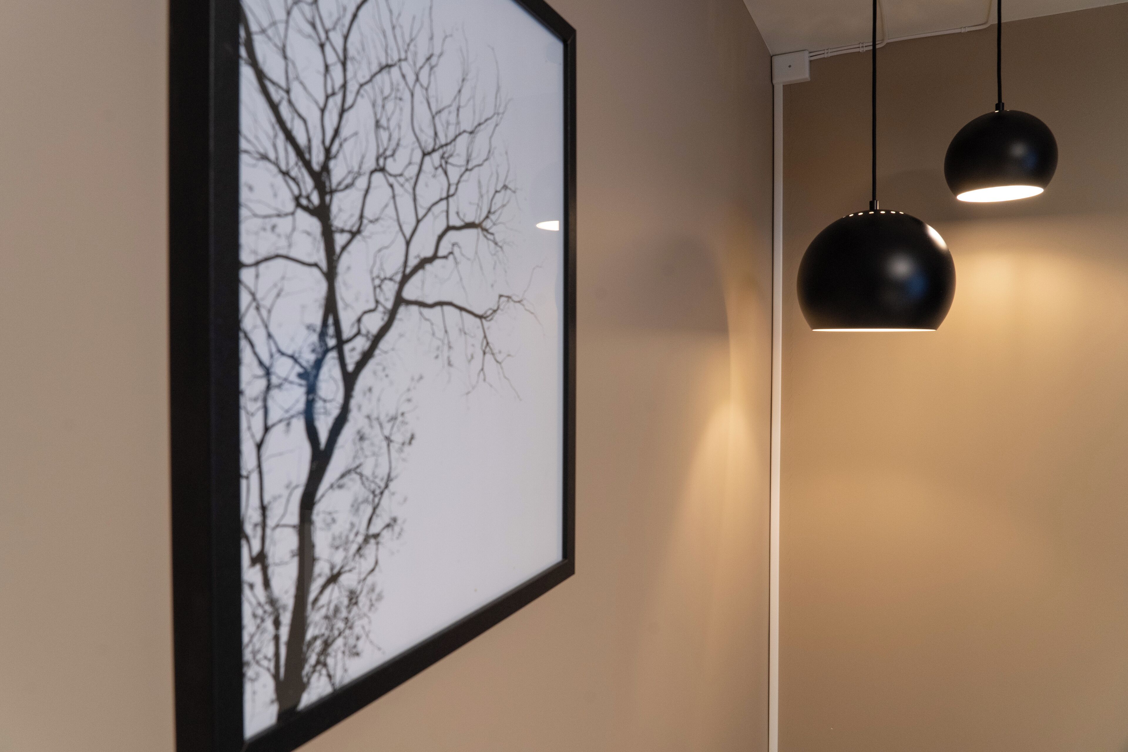 Um quadro com uma árvore sem folhas na parede, duas luminárias pendentes pretas e uma parede lisa.