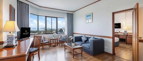 Quarto de hotel com piso de madeira, um sofá, uma televisão, uma escrivaninha e vista para a cidade.