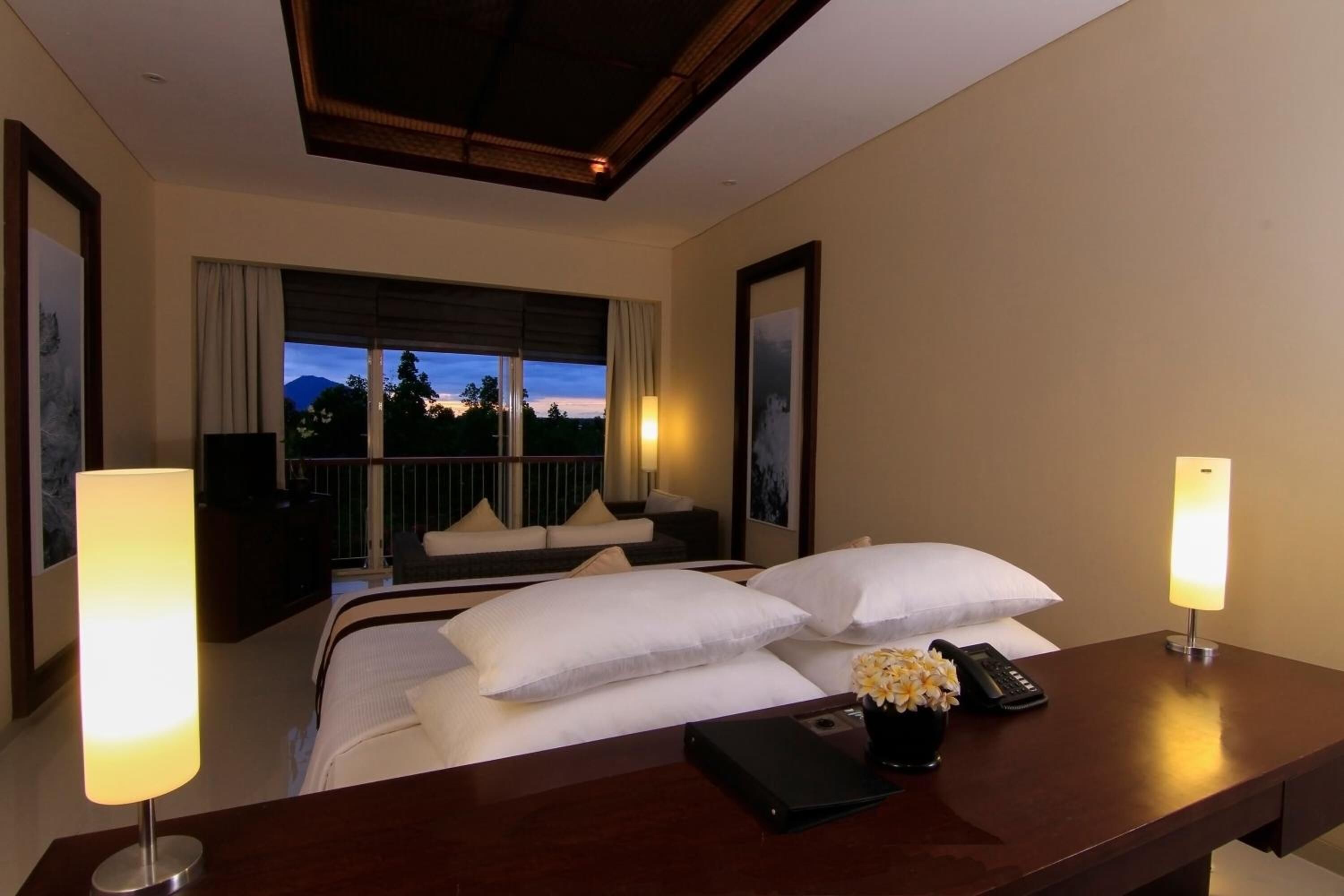 Quarto de hotel com cama, abajures, escrivaninha com telefone e um vaso de flores, além de vista para as montanhas pela janela.