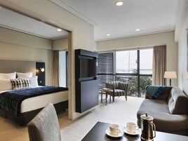 Quarto de hotel com cama, televisão, sofá, mesa com chávenas e cafeteira, e vista da cidade através de grandes janelas.
