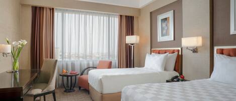 Quarto de hotel com duas camas, uma escrivaninha, uma cadeira, uma mesa pequena e vista da cidade através de janelas amplas.