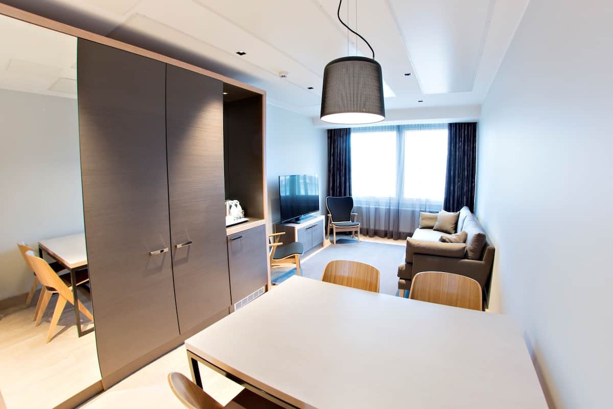 Quarto de hotel moderno com área de refeições, sofá, televisão e uma janela grande com cortinas.