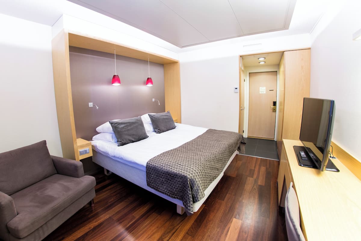 Quarto de hotel moderno com cama, escrivaninha com computador, sofá e duas luminárias pendentes.