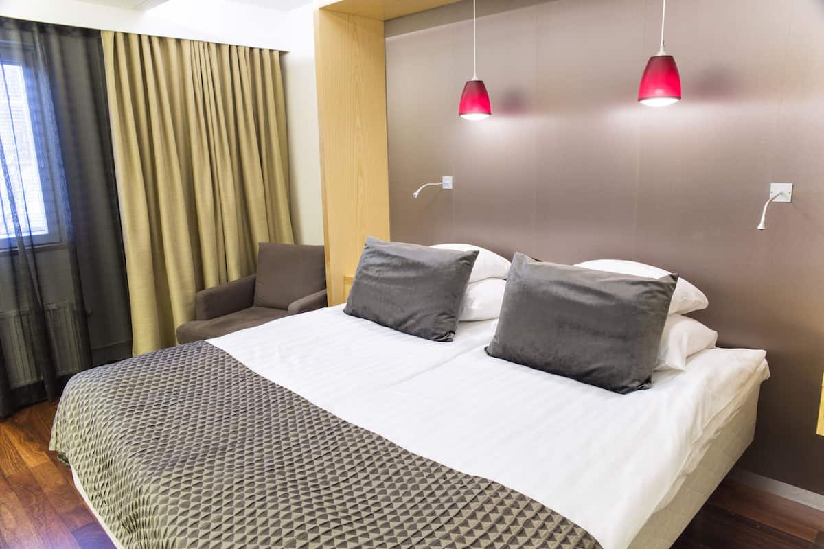 Quarto de hotel moderno com uma cama grande, duas luminárias de cabeceira, um sofá e vista para a cidade.