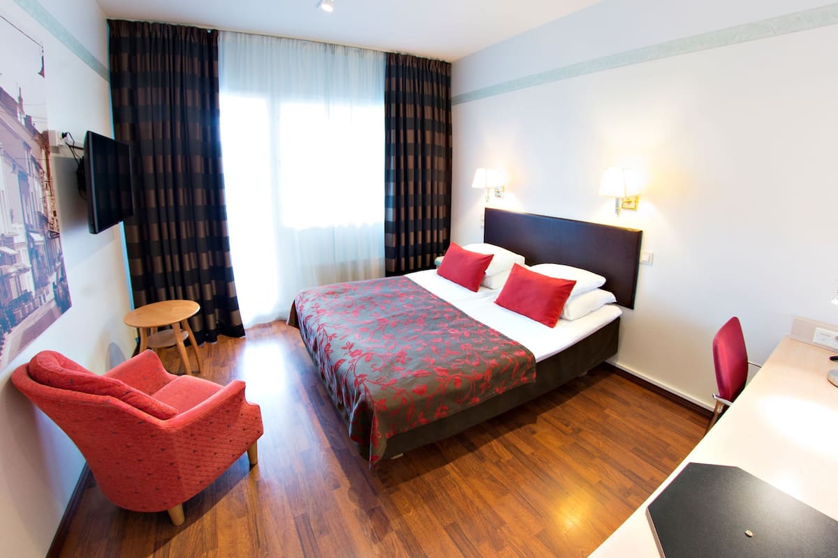 Quarto de hotel com uma cama, uma poltrona vermelha, uma mesinha e uma escrivaninha.