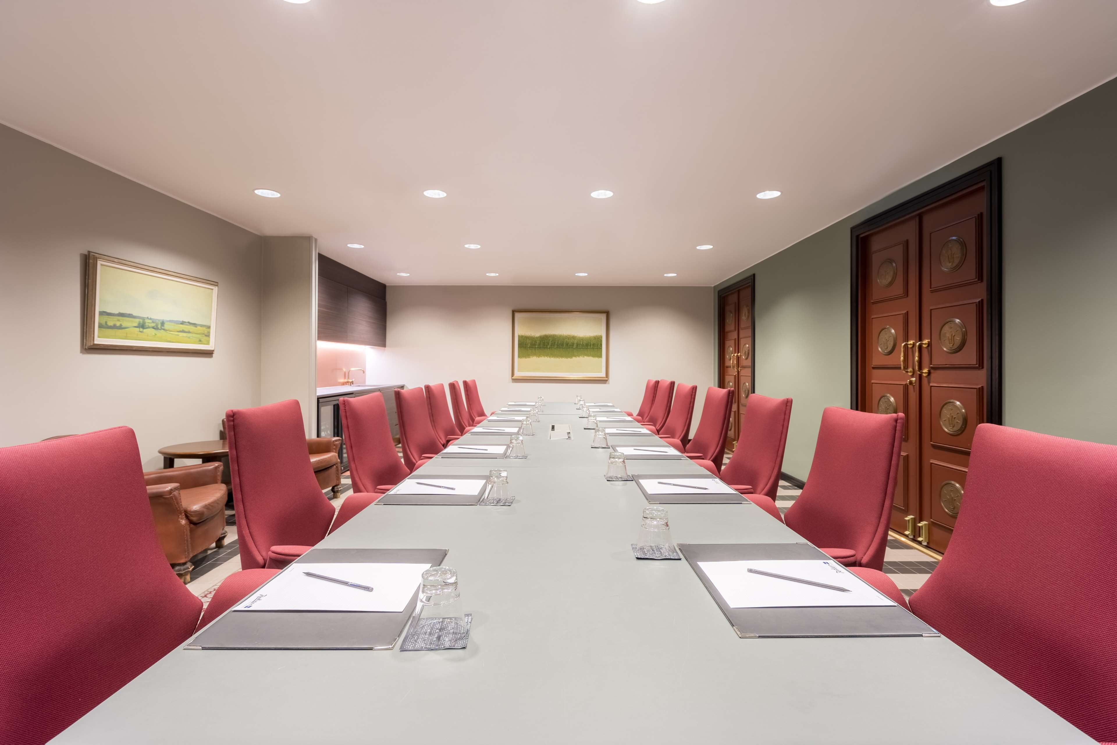Uma sala de conferência com uma mesa comprida, cadeiras vermelhas e uma porta de madeira com um relógio.