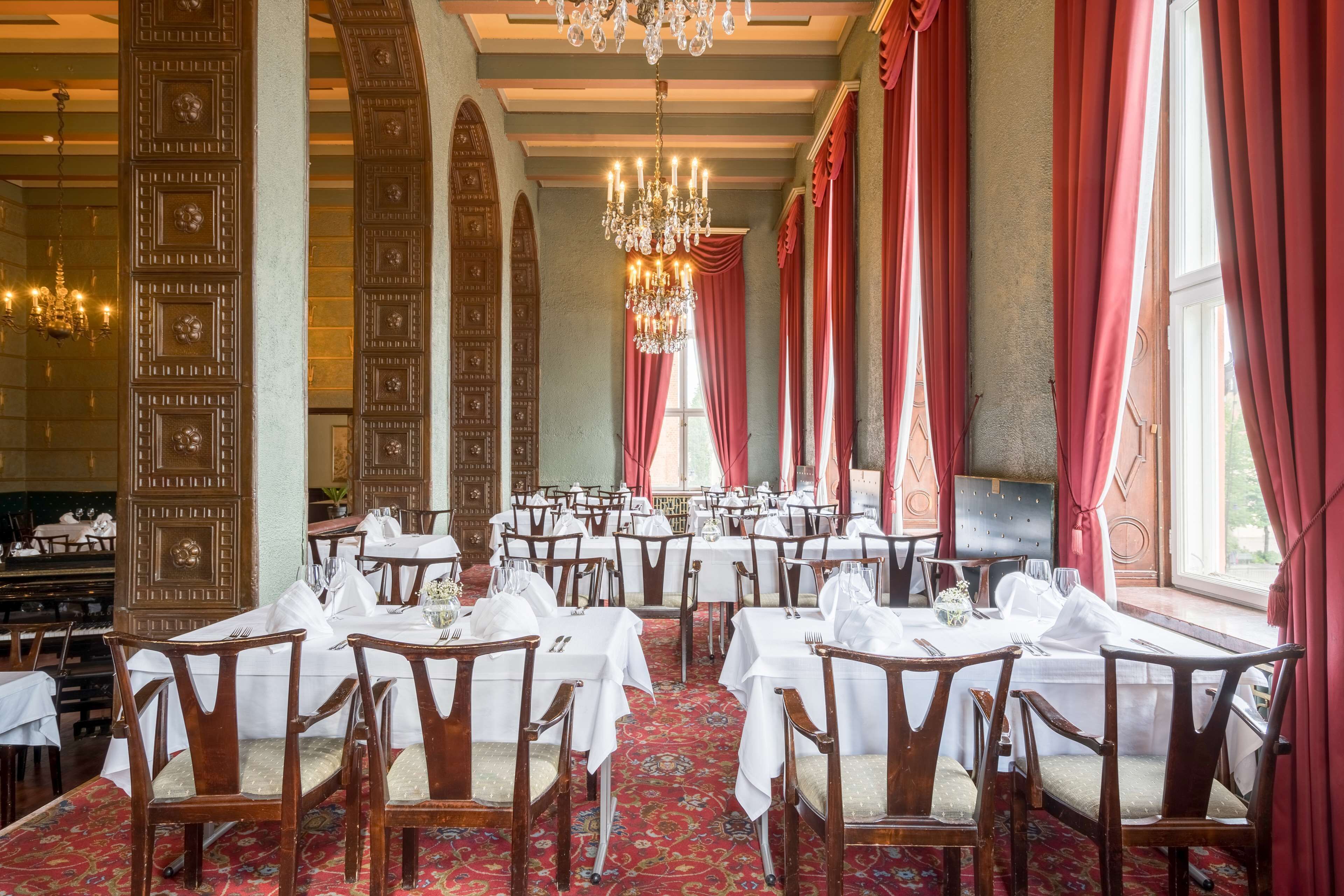 Uma sala de jantar formal com várias mesas preparadas para o serviço, painéis de madeira ornamentados e um candelabro.