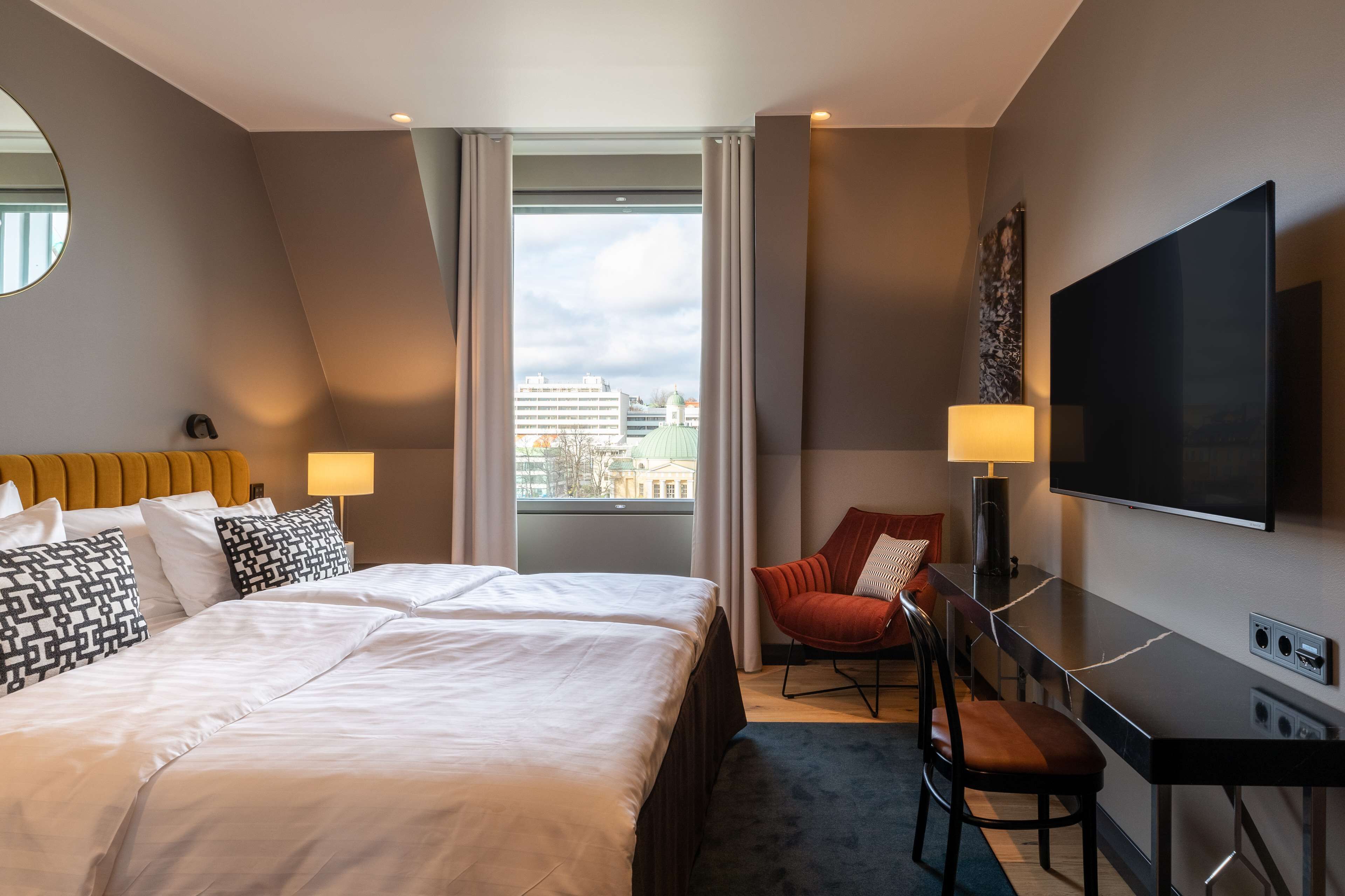 Quarto de hotel com uma cama grande, uma escrivaninha com televisão e vista para a cidade pela janela.