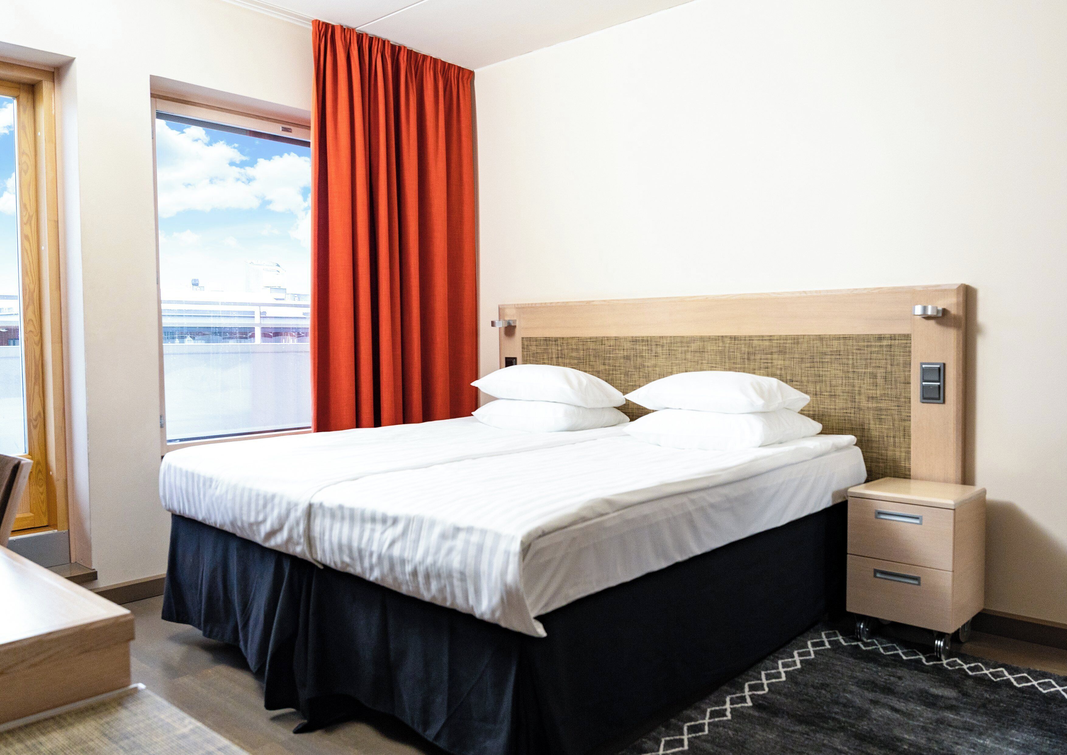 Quarto de hotel com cama, mesa de cabeceira, gaveta, janela com cortinas e vista para o exterior.