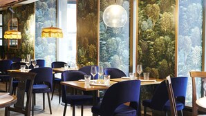 Restaurante com mesas de madeira e cadeiras azuis, um espelho grande e uma parede com um mural de temática natural.