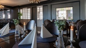 Uma sala de jantar com mesa de madeira escura posta com guardanapos brancos, uma vela branca e um vaso de vidro com folhagem.