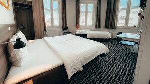 Quarto de hotel com uma cama grande, uma cama menor, uma escrivaninha e uma cadeira. Há janelas com cortinas, um radiador e uma televisão montada na parede.