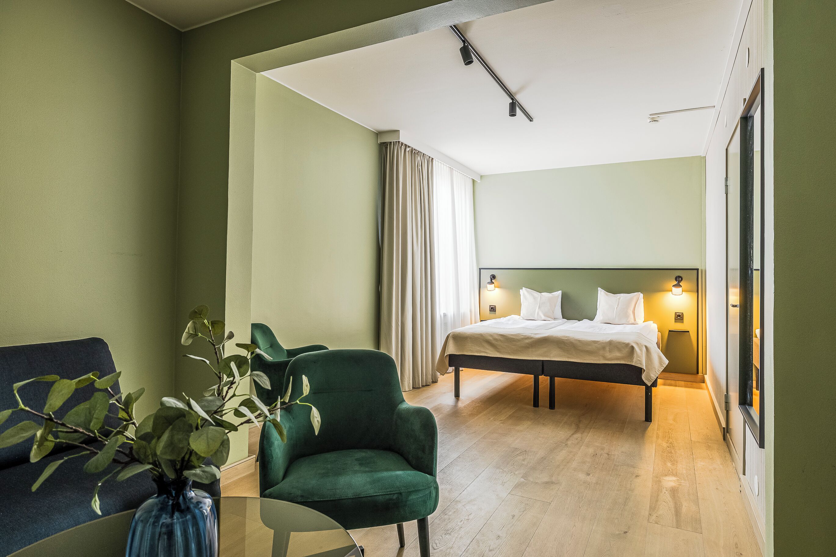 Quarto de hotel com uma cama, uma poltrona verde, uma mesa pequena com um vaso e uma janela com cortinas.