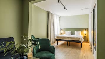 Quarto de hotel com uma cama, uma poltrona verde, uma mesa pequena com um vaso e uma janela com cortinas.