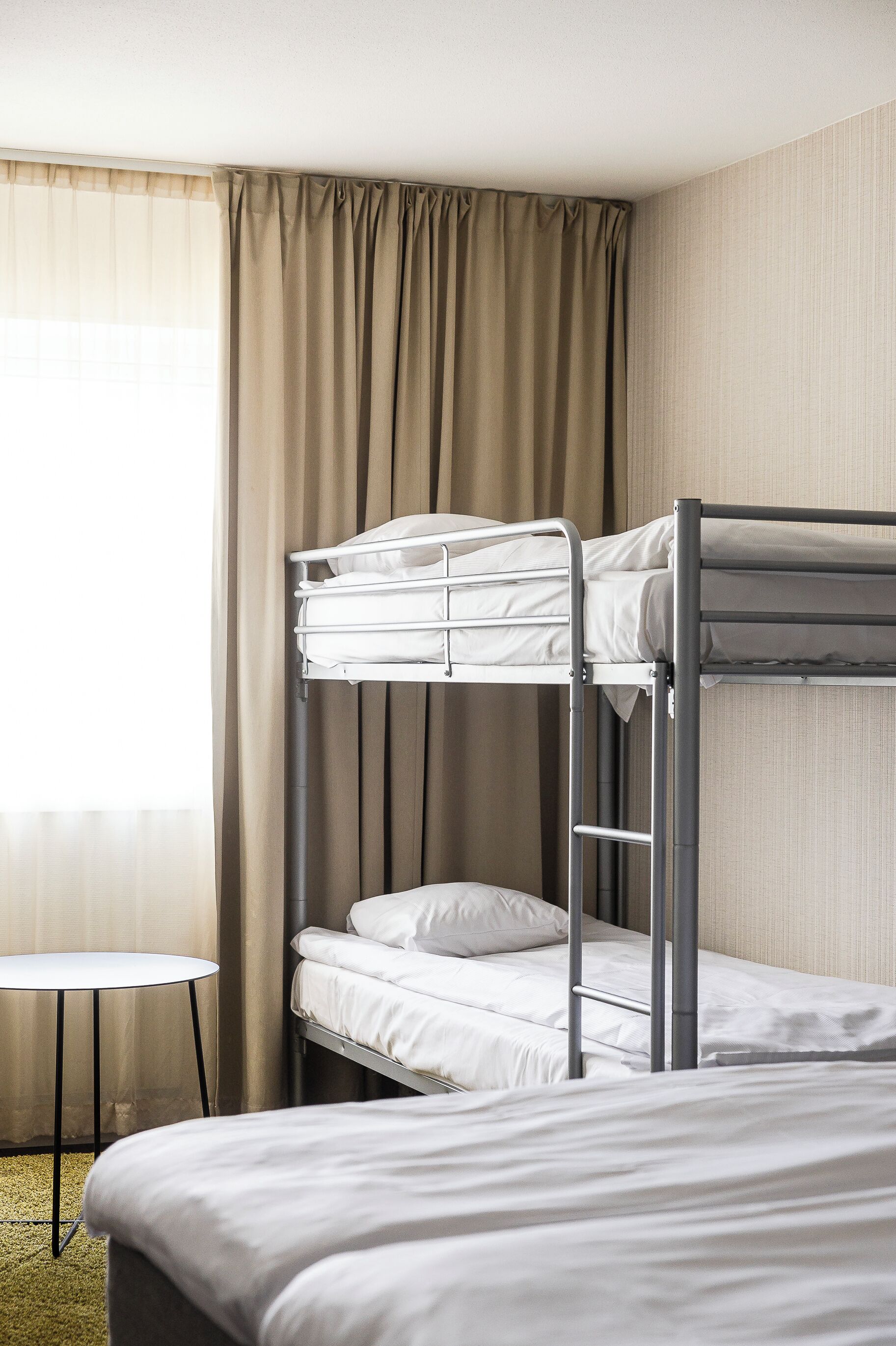 Uma cama beliche com escada, uma mesinha redonda e uma janela com cortinas.